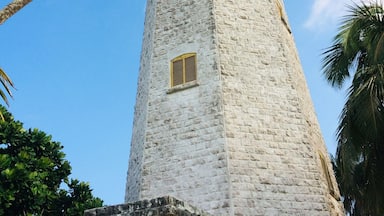 Um farol alto e branco de pedra, com duas janelas, rodeado por palmeiras e um céu azul límpido.