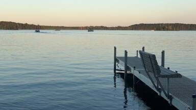 Um lago calmo com um píer que se estende sobre a água, cercado por árvores e um céu claro.