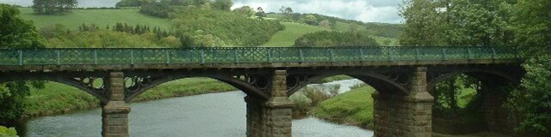Uma ponte de pedra sobre um rio, com grades verdes, cercada por vegetação exuberante e colinas.