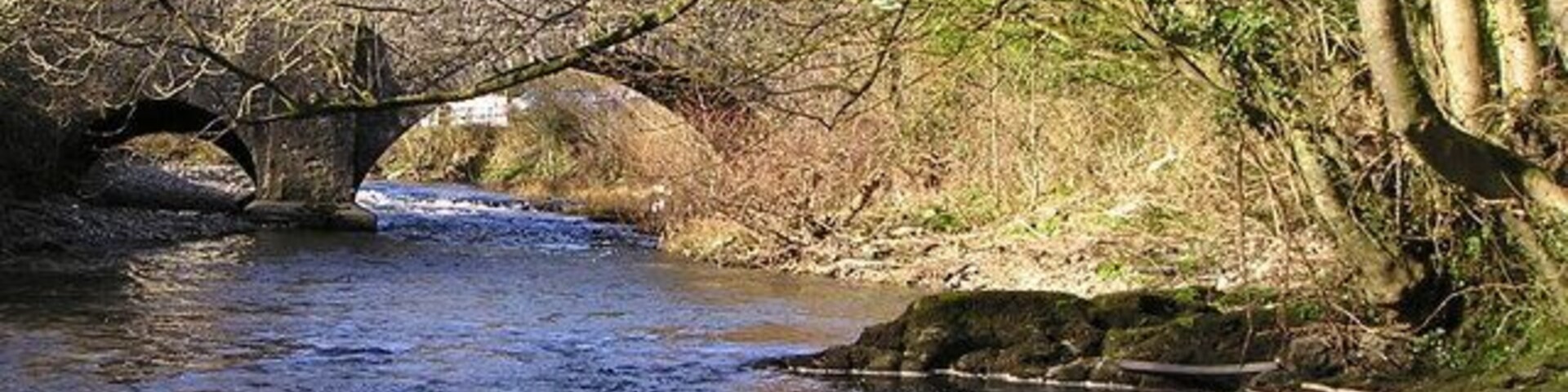 Uma ponte de pedra sobre um rio, com árvores e pedras ao redor.