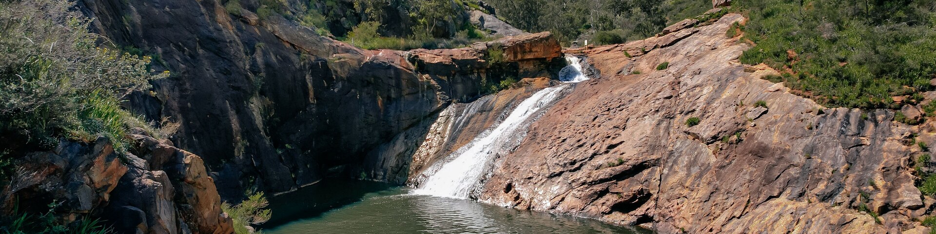 Uma cascata deságua num rio, rodeado por terreno rochoso e vegetação.