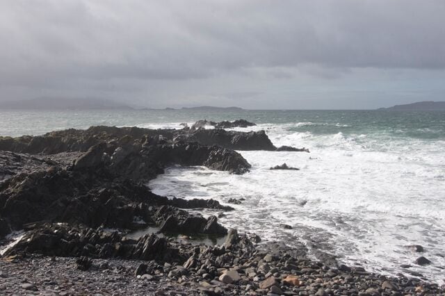 Uma costa rochosa com ondas quebrando nas pedras sob um céu nublado.
