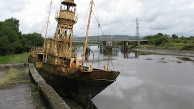 Um barco antigo e enferrujado atracado na margem de um rio, com uma ponte ao fundo.