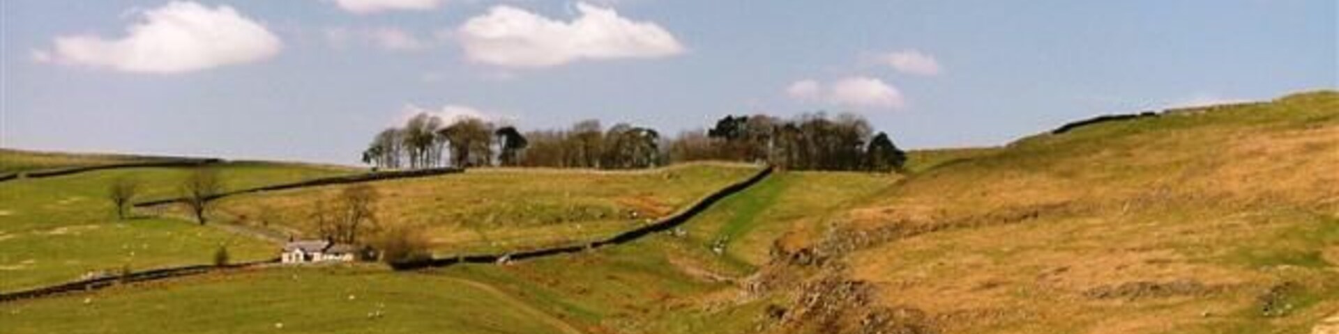 Paisagem rural com colinas ondulantes, um muro de pedra e um pequeno edifício branco.