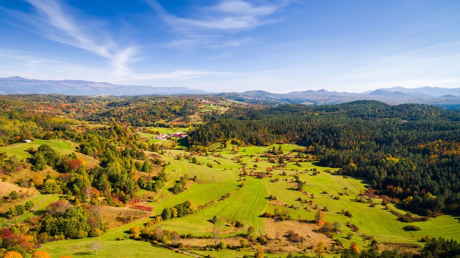 Vista panorâmica de uma paisagem montanhosa com campos verdes ondulantes, florestas densas e cadeias de montanhas distantes sob um céu azul límpido.