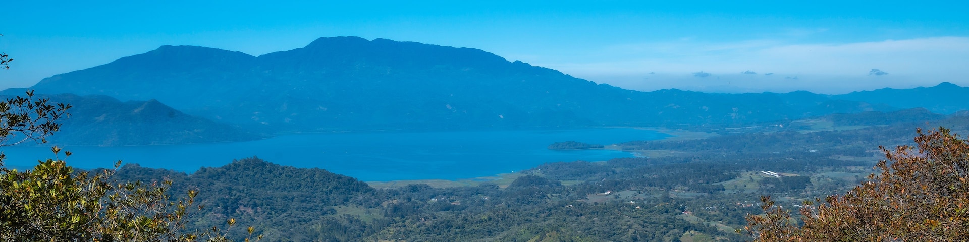 Paisagem montanhosa com céu azul límpido, uma grande massa de água e vegetação verde exuberante em primeiro plano.