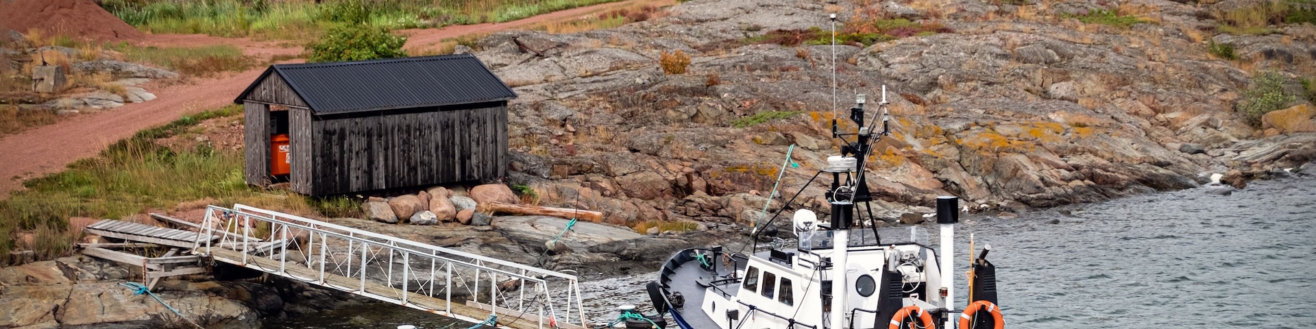 Um barco azul atracado numa costa rochosa com um cais de madeira, um pequeno armazém e uma área verde ao fundo.