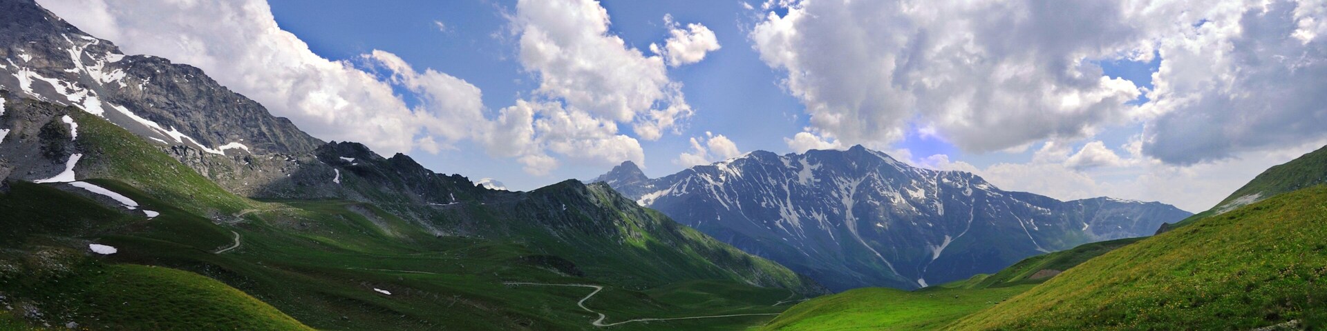 Paisagem montanhosa com vales verdes e picos nevados sob um céu parcialmente nublado.