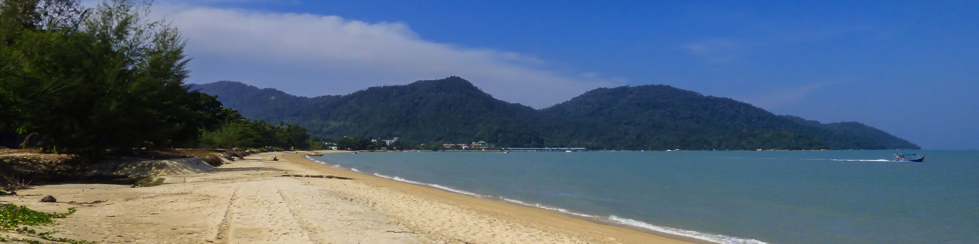 Uma praia arenosa com águas azuis cristalinas, um barco ao longe e montanhas ao fundo.