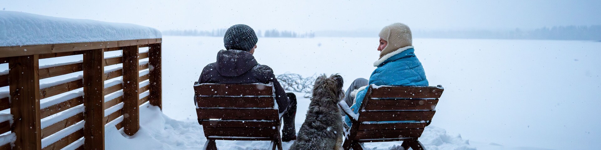 Duas pessoas sentadas em bancos de madeira em uma paisagem nevada, com uma cerca de madeira e um cachorro.