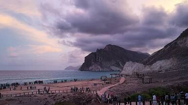 Paisagem costeira com um penhasco rochoso, uma praia com pessoas e céu claro.