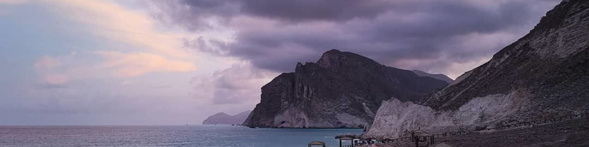 Paisagem costeira com um penhasco rochoso, uma praia com pessoas e céu claro.