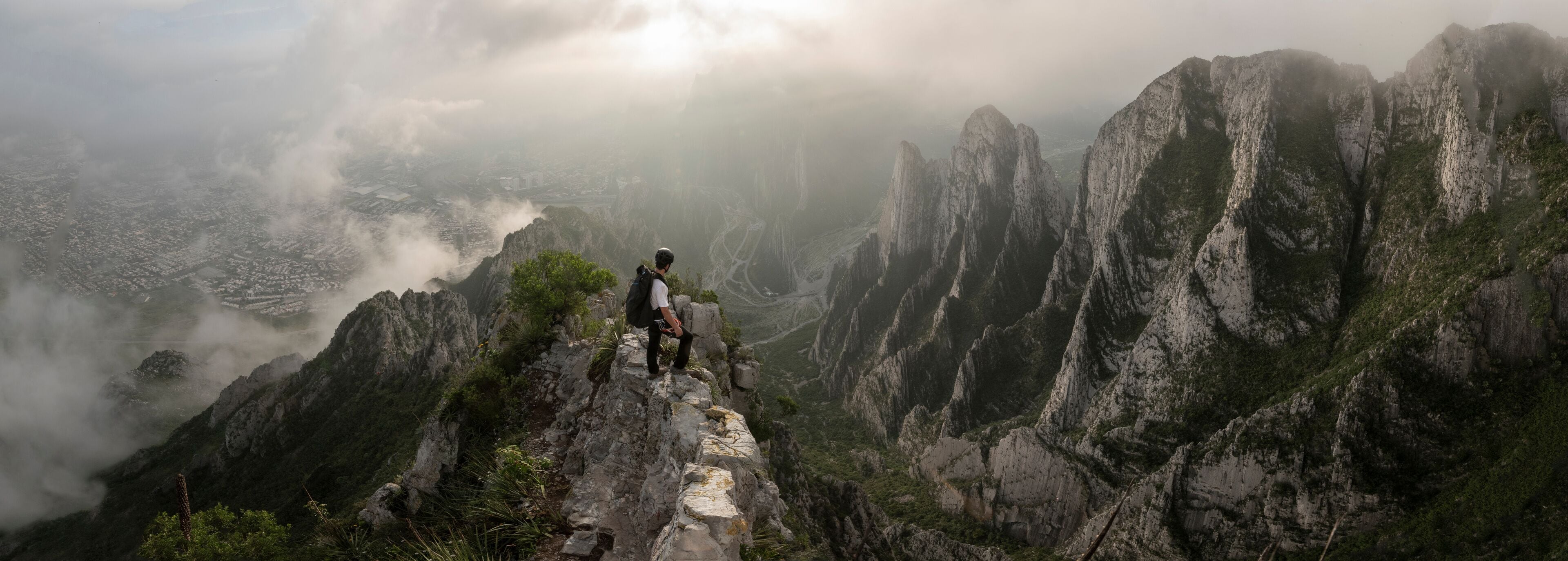 Uma pessoa está em cima de um cume de montanha, com penhascos íngremes e um vale lá embaixo.