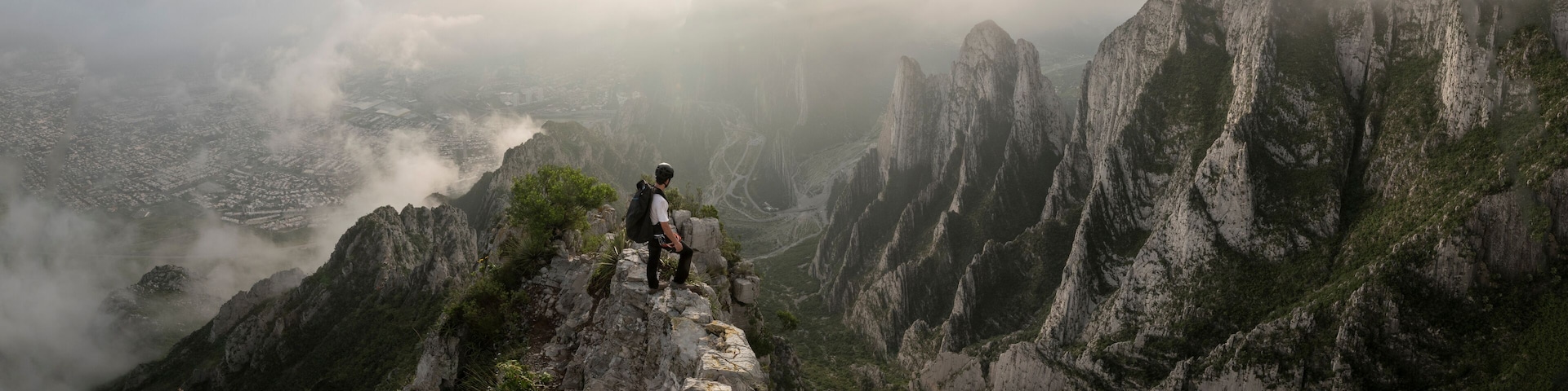 Uma pessoa está em cima de um cume de montanha, com penhascos íngremes e um vale lá embaixo.