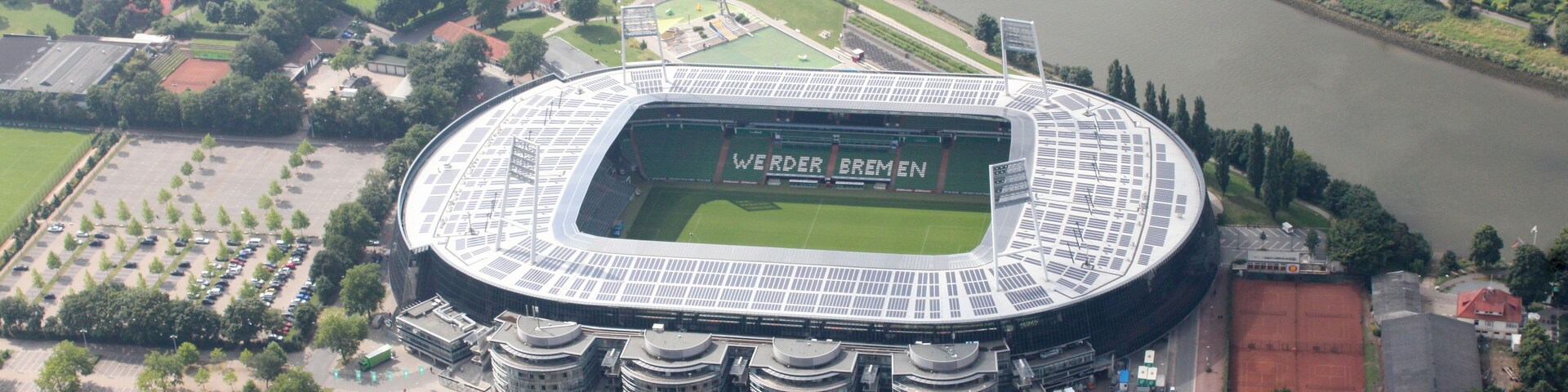 Vista aérea de um estádio com 'WERDER BREMEN' escrito no campo, cercado por edifícios residenciais e um rio.