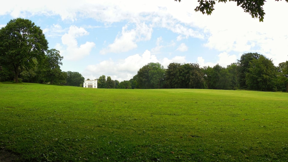 Um campo gramado com um edifício branco ao longe, cercado por árvores.
