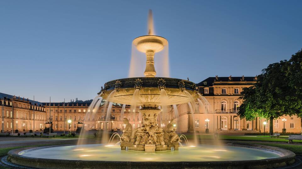 Uma grande fonte iluminada com vários jatos de água em frente a um edifício histórico ao anoitecer.