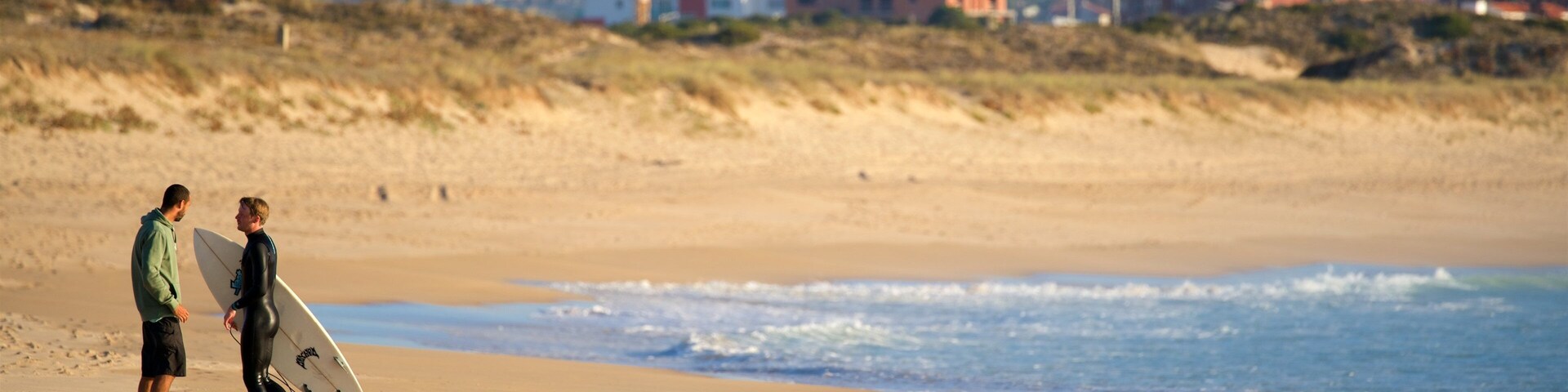 Duas pessoas caminham numa praia arenosa com ondas do mar, uma turbina eólica e edifícios ao fundo.