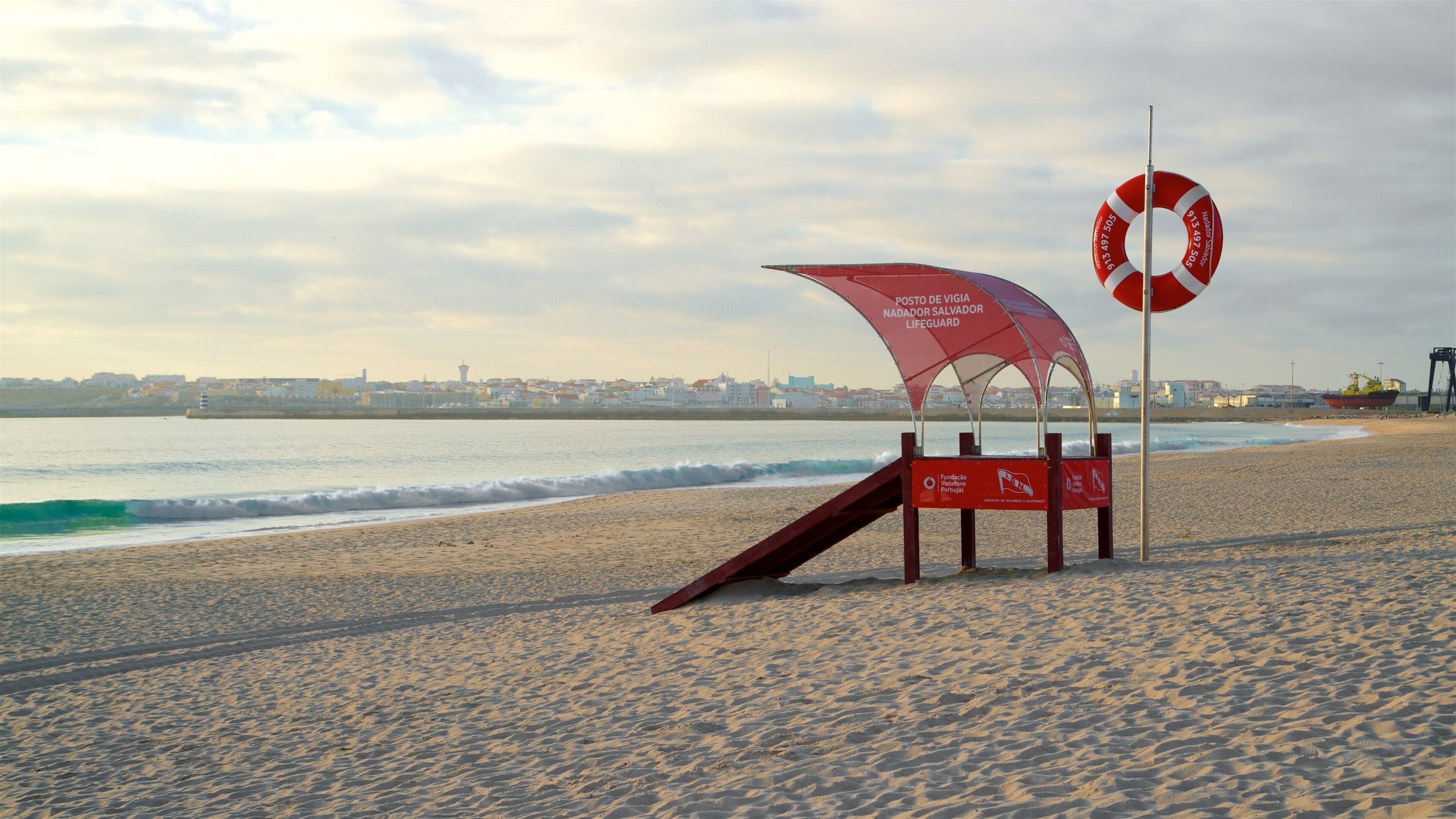 Um posto de salva-vidas com um guarda-sol vermelho numa praia com ondas, um flutuador e uma paisagem urbana ao fundo.