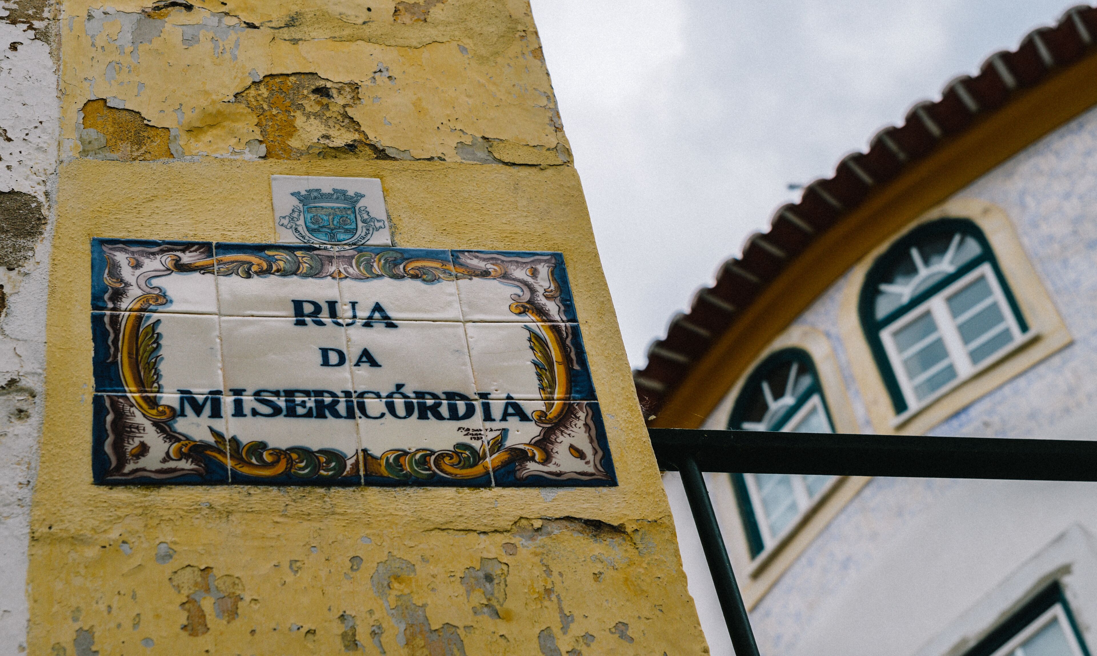 Uma placa de rua desgastada com 'RUA DA MISERICÓRDIA' em uma parede amarela, um brasão acima dela e um edifício com janelas em arco ao fundo.