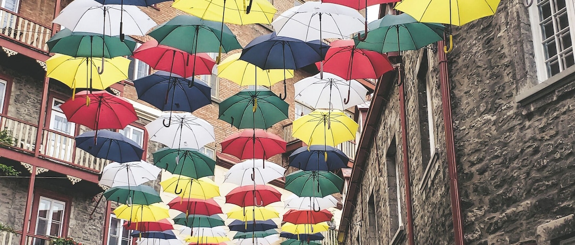 Uma rua com guarda-sóis coloridos pendurados, edifícios e pessoas caminhando.