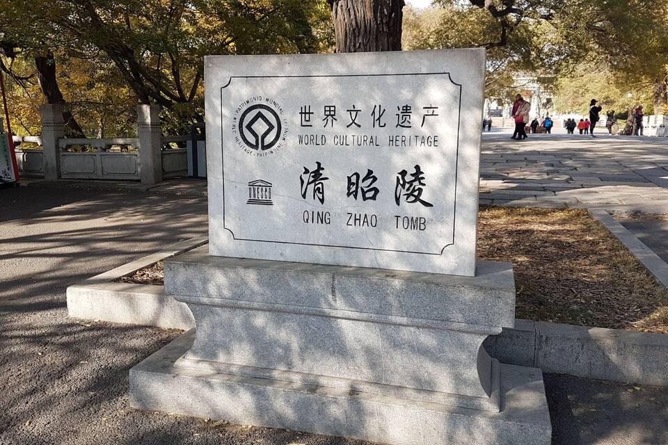 Um monumento de pedra com inscrições em chinês e inglês, cercado por árvores e uma área pavimentada, com pessoas ao fundo.