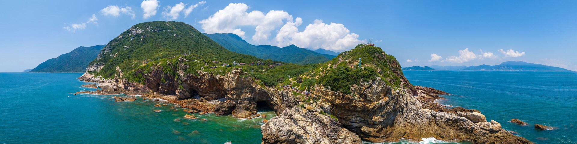 Paisagem costeira com falésias rochosas, um barco na água e montanhas ao longe sob um céu claro.