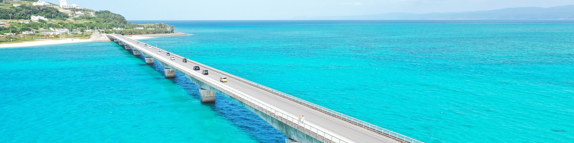 Uma longa ponte sobre águas turquesa, com uma vila costeira e uma montanha ao fundo.