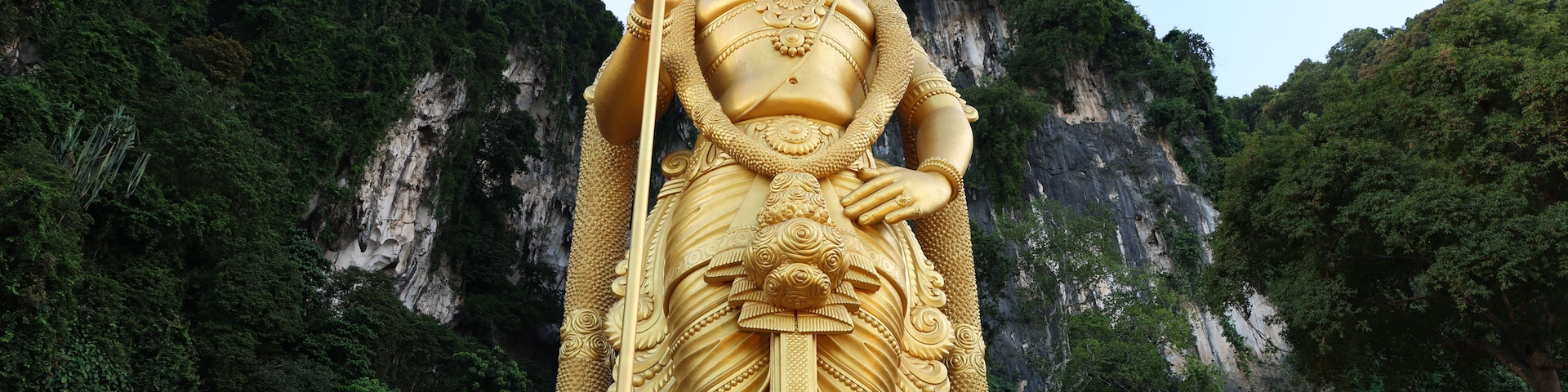 Grande estátua dourada de uma divindade segurando um cajado, com penhascos verdes exuberantes ao fundo.