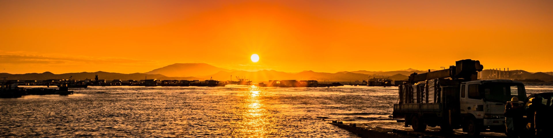 Um pôr do sol sobre a água, com um caminhão e estruturas industriais em primeiro plano.