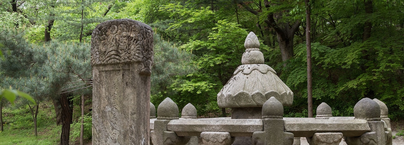 Um monumento de pedra com esculturas detalhadas, rodeado por uma cerca tradicional e situado numa floresta verdejante.