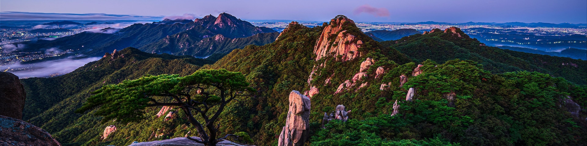 Paisagem montanhosa com um pico rochoso imponente, vegetação exuberante e um horizonte urbano ao longe.