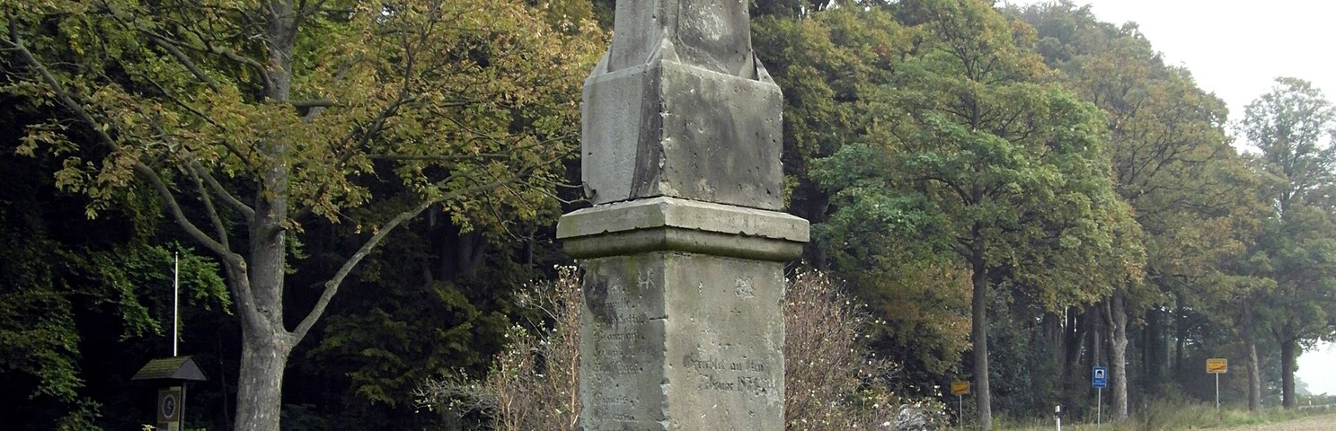 Um monumento de pedra com uma cruz no topo, rodeado por relva e árvores.