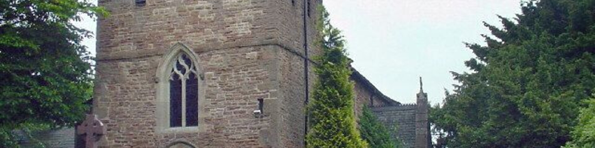 Uma torre de pedra histórica com uma janela em arco pontiagudo e um pequeno campanário ao fundo.