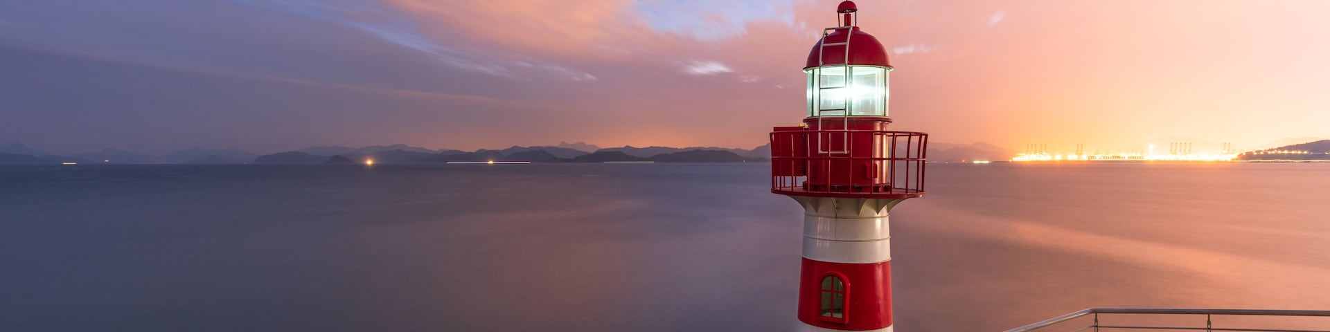 Um farol vermelho e branco numa formação rochosa, com um mar calmo e um céu ao pôr do sol.