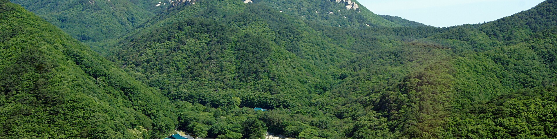 Paisagem montanhosa com uma grande formação rochosa ao fundo, um templo com uma estátua em primeiro plano e um edifício com telhado verde aninhado entre as árvores.