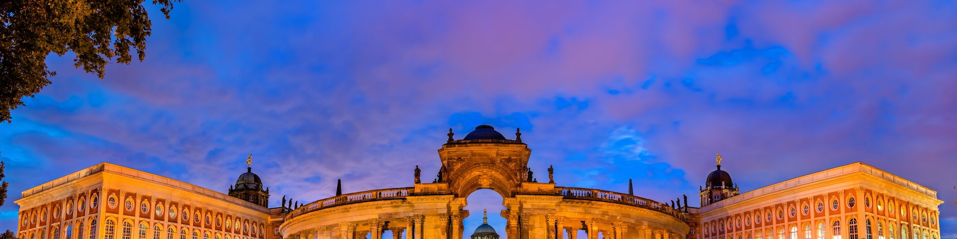 Um imponente edifício histórico com um arco central, ladeado por duas alas, sob um céu crepuscular.