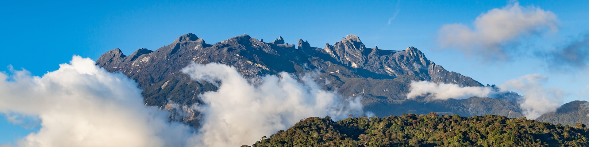 Cordilheira com nuvens na base, floresta densa e céu azul límpido.