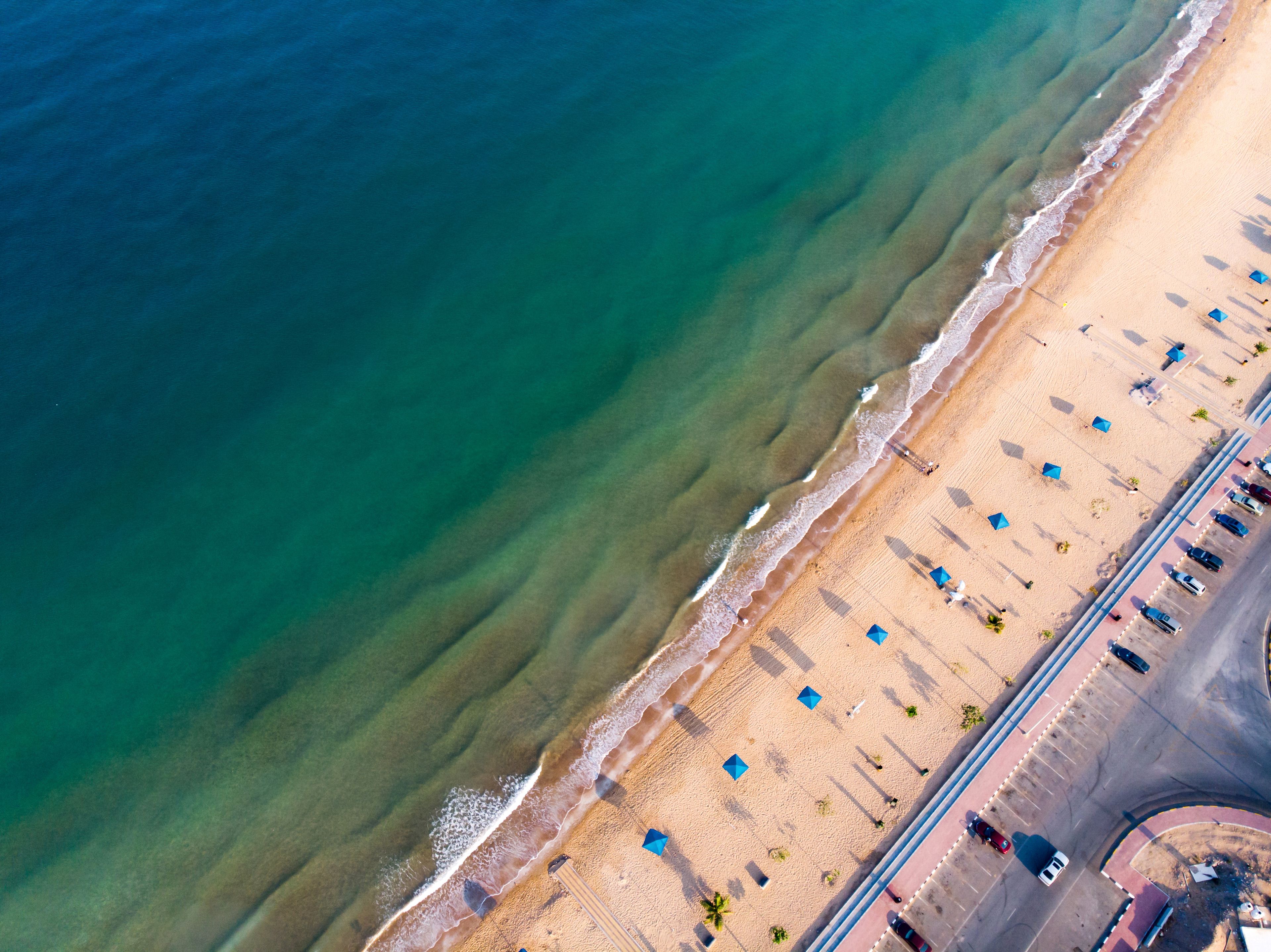 Vista aérea de uma praia com águas azuis cristalinas, areal dourado e estacionamento com guarda-sóis azuis.