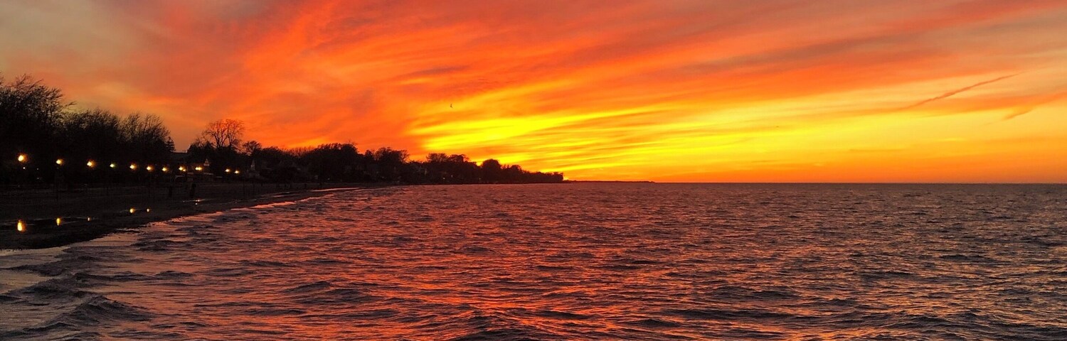 Pôr do sol sobre a água, com silhueta da linha costeira e postes de luz.