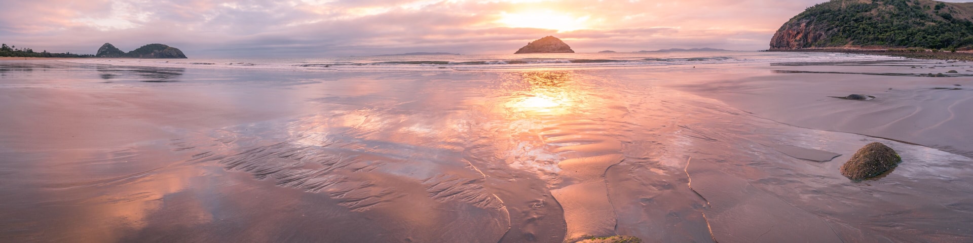 Uma praia com águas calmas, uma rocha grande em primeiro plano e um pôr do sol ao fundo.