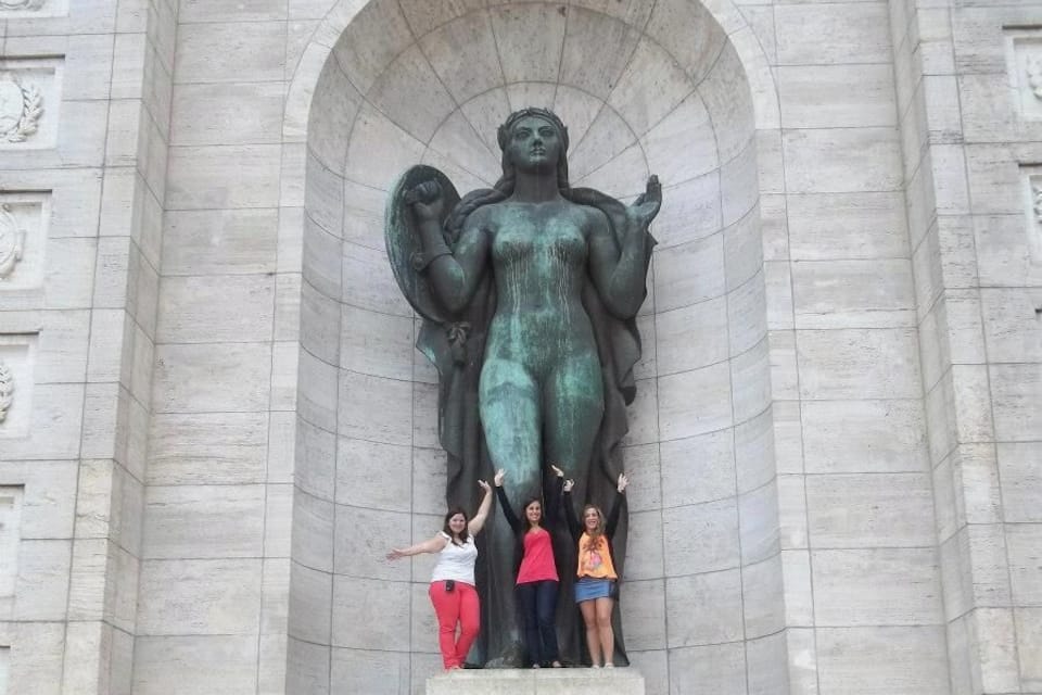 Uma grande estátua de bronze de uma figura alada sobre um pedestal, em frente a uma fachada de construção de pedra com detalhes arquitetônicos clássicos.