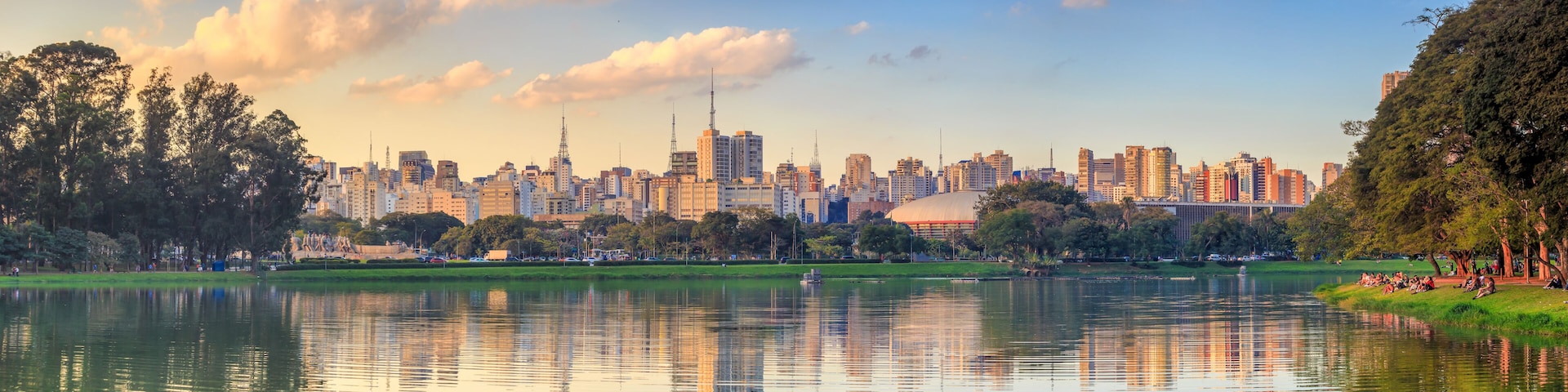Silhueta urbana com um estádio grande, um lago refletindo edifícios e árvores na margem.