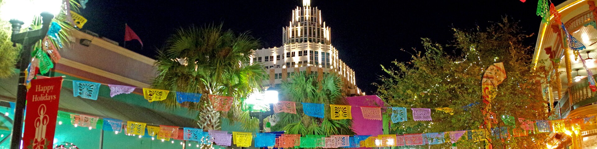 Uma rua bem iluminada com bandeiras coloridas, um edifício com torre e placas indicando restaurantes.