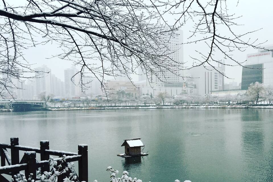 Paisagem nevada com um cais de madeira e uma pequena casa flutuante em um rio calmo, cercada por árvores cobertas de neve e edifícios ao fundo.