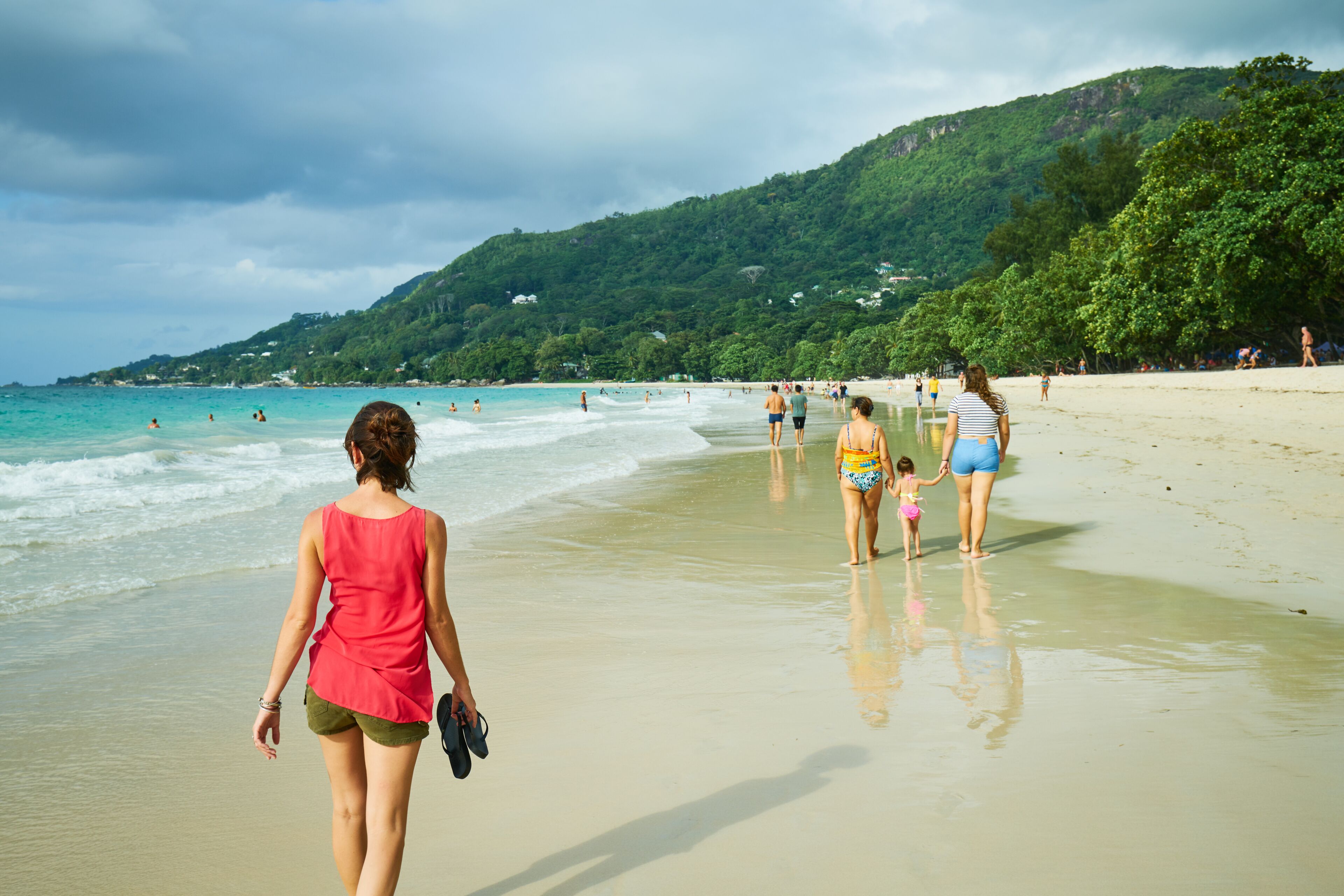 Uma praia com pessoas caminhando pela costa, céu claro e uma montanha ao fundo.