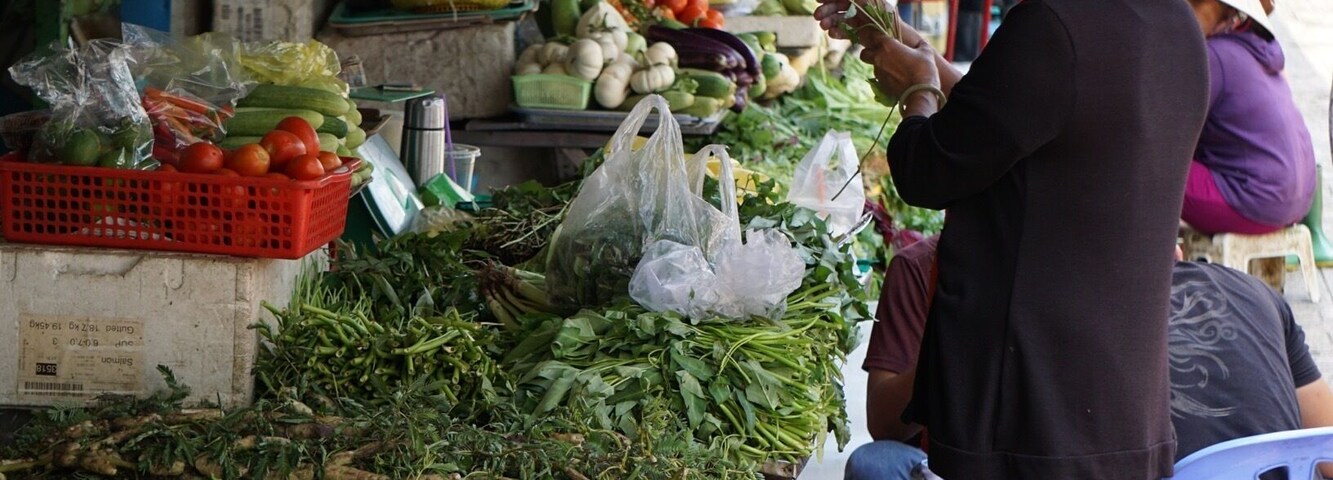 Um mercado ao ar livre movimentado, com diversos legumes e frutas frescos expostos em mesas e cestas.