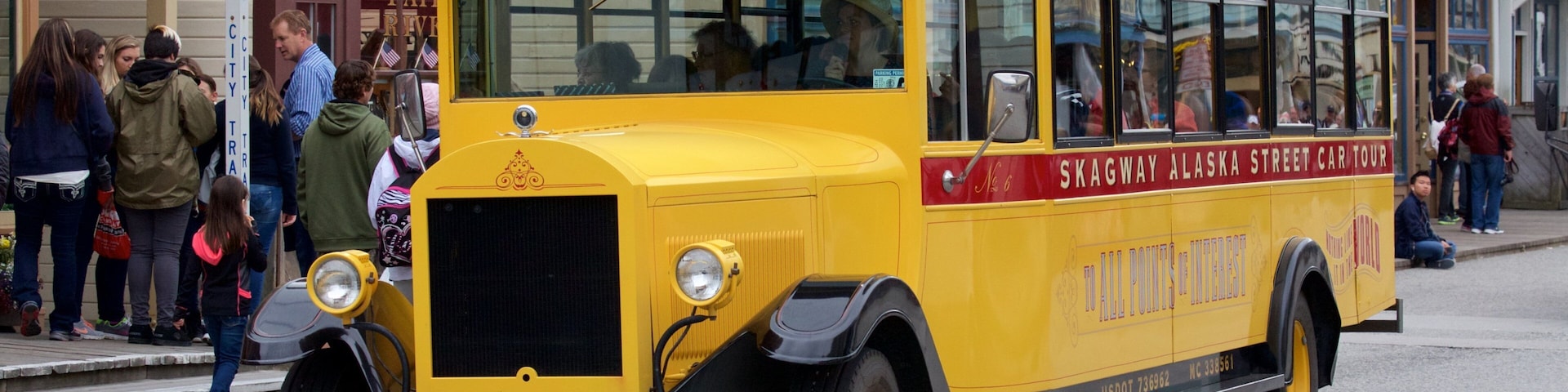 Um elétrico antigo amarelo com a inscrição "Skagway Alaska Street Car Tour" na lateral, estacionado numa rua com pessoas por perto.