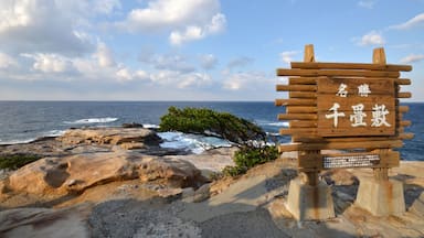 Uma placa de madeira com caracteres chineses, uma orla rochosa e o oceano ao fundo.
