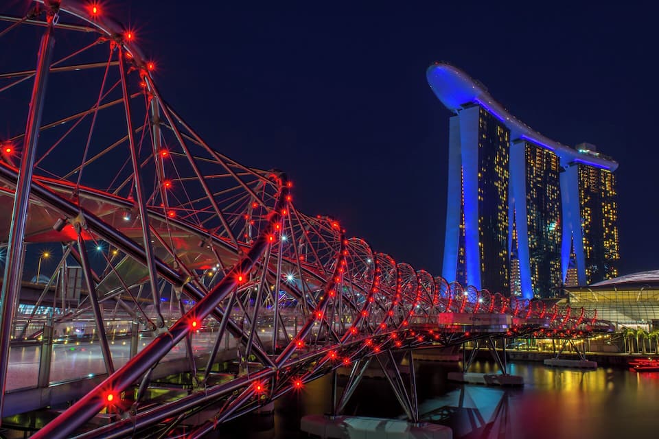 Vista noturna de uma grande roda gigante com luzes vermelhas, uma ponte moderna e o hotel Marina Bay Sands com luzes azuis ao fundo.